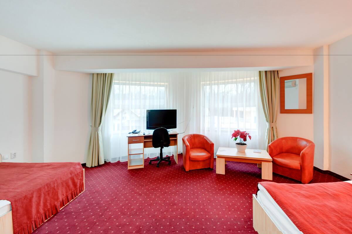 Quarto de hotel com duas camas de solteiro, uma escrivaninha com computador e cadeira, uma mesinha com um vaso, e duas poltronas.