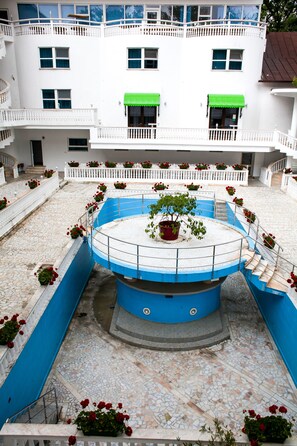 Pátio com uma fonte central, rodeado por canteiros de flores e um edifício branco com varandas.