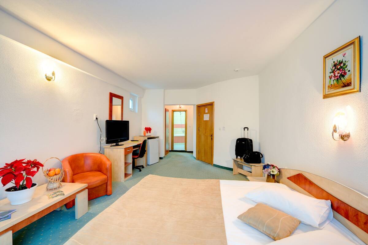 Quarto de hotel com cama, escrivaninha, cadeira, televisão e porta para outro aposento.
