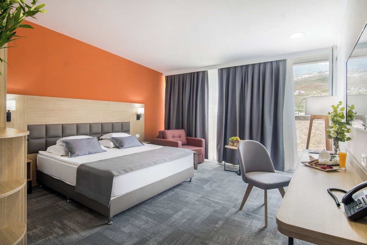 Quarto de hotel com uma cama, uma escrivaninha, uma cadeira, uma pequena mesa com uma planta e vista para o exterior.
