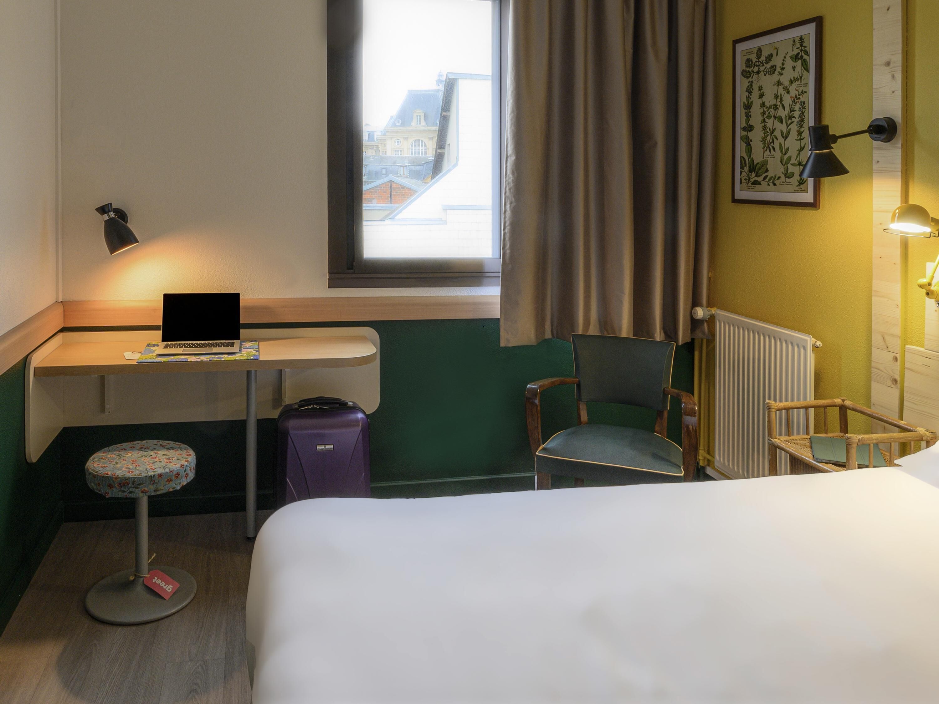 Quarto de hotel com cama, escrivaninha, um laptop, cadeira, mala e janela com cortinas.