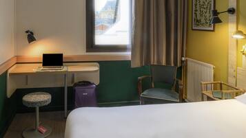 Quarto de hotel com cama, escrivaninha, um laptop, cadeira, mala e janela com cortinas.