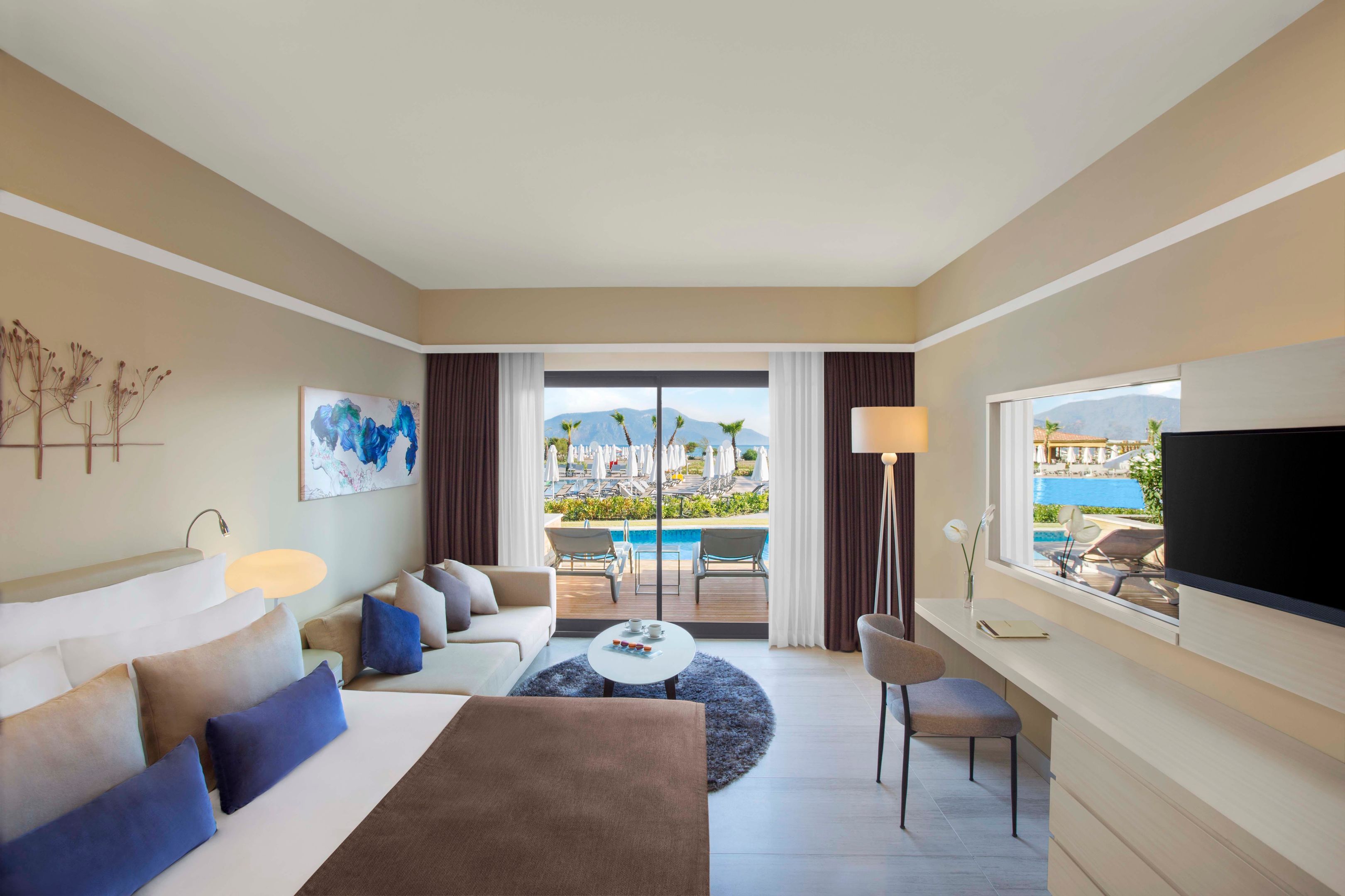 Quarto de hotel com cama, sofá, televisão e vista para uma piscina e montanhas.