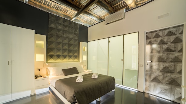 Quarto de hotel moderno com uma cama grande, uma mesinha de cabeceira, parede com padrões geométricos e uma porta deslizante com texto informativo.
