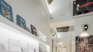 Um corredor com paredes brancas, estantes exibindo livros e quadros, e um grande mural na parede.