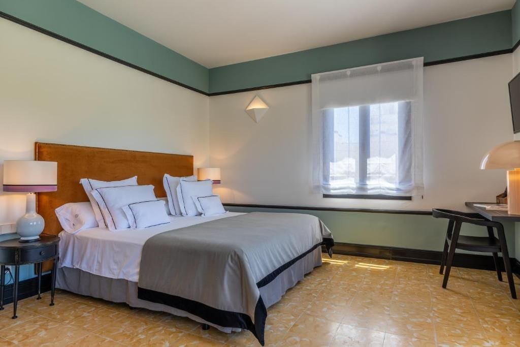 Quarto de hotel com cama, mesas de cabeceira, janela com cortinas e escrivaninha com cadeira.