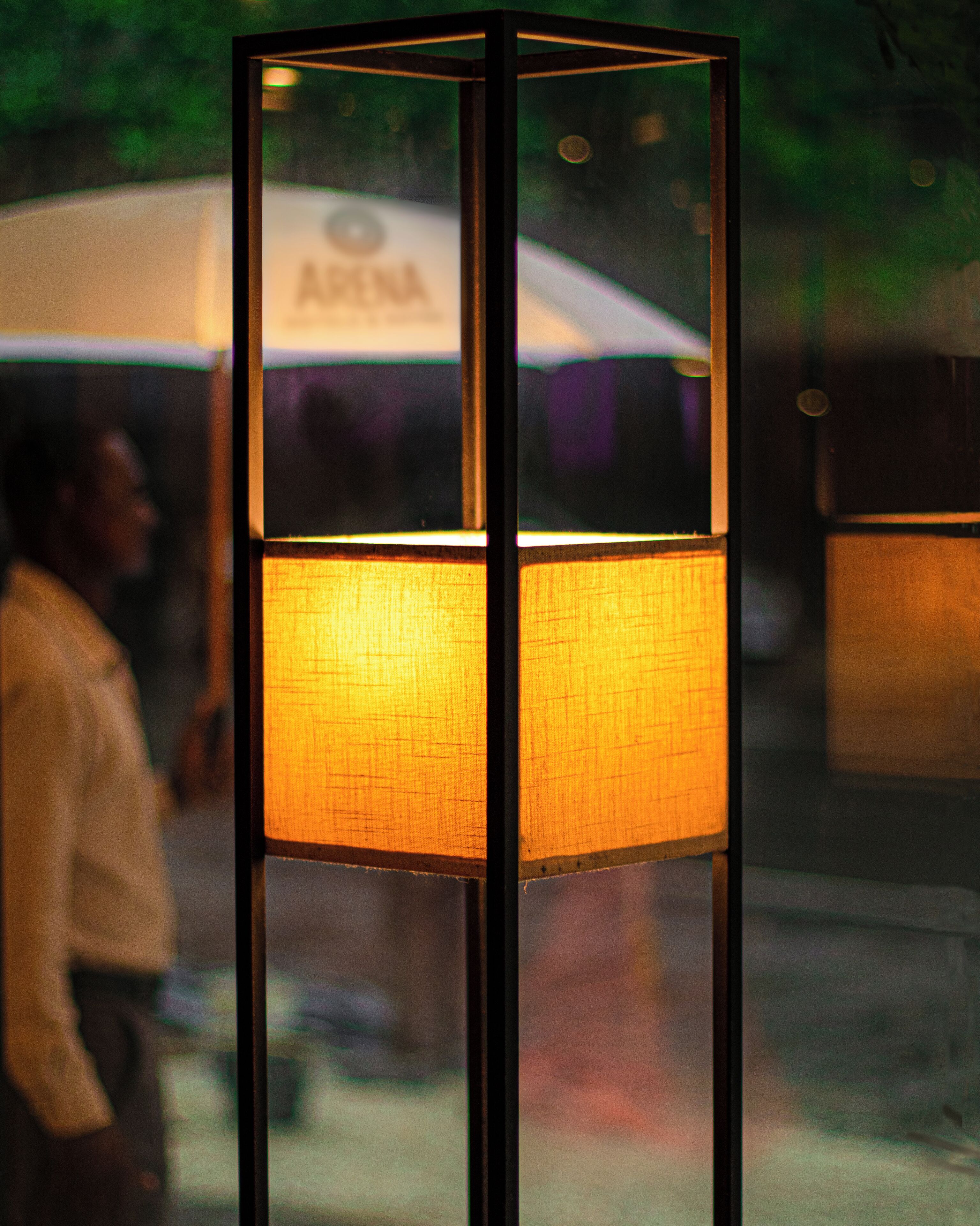 Luminária alta e retangular com estrutura preta e interior iluminado, em frente a um fundo desfocado onde se vê uma pessoa.