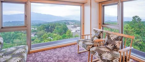Quarto com grandes janelas oferecendo vista para uma paisagem montanhosa. Há uma cadeira de madeira com almofada estampada em folha e uma mesa pequena.