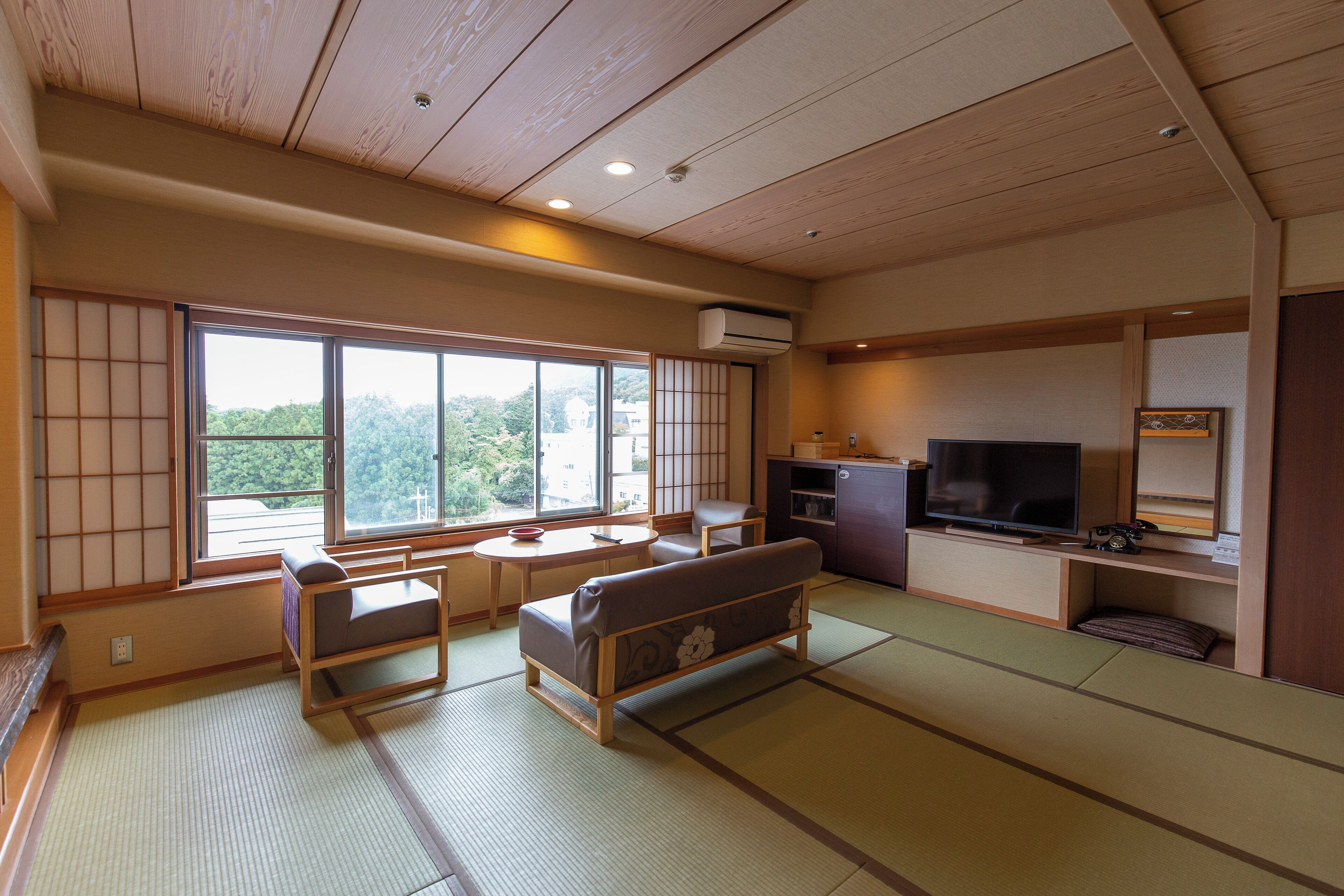 Quarto tradicional japonês com piso de tatame, mesa baixa de tatame, sofá de madeira rústico, televisão e uma porta de correr que dá para uma varanda com vista para o verde.