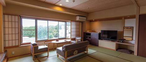 Quarto tradicional japonês com piso de tatame, mesa baixa de tatame, sofá de madeira rústico, televisão e uma porta de correr que dá para uma varanda com vista para o verde.