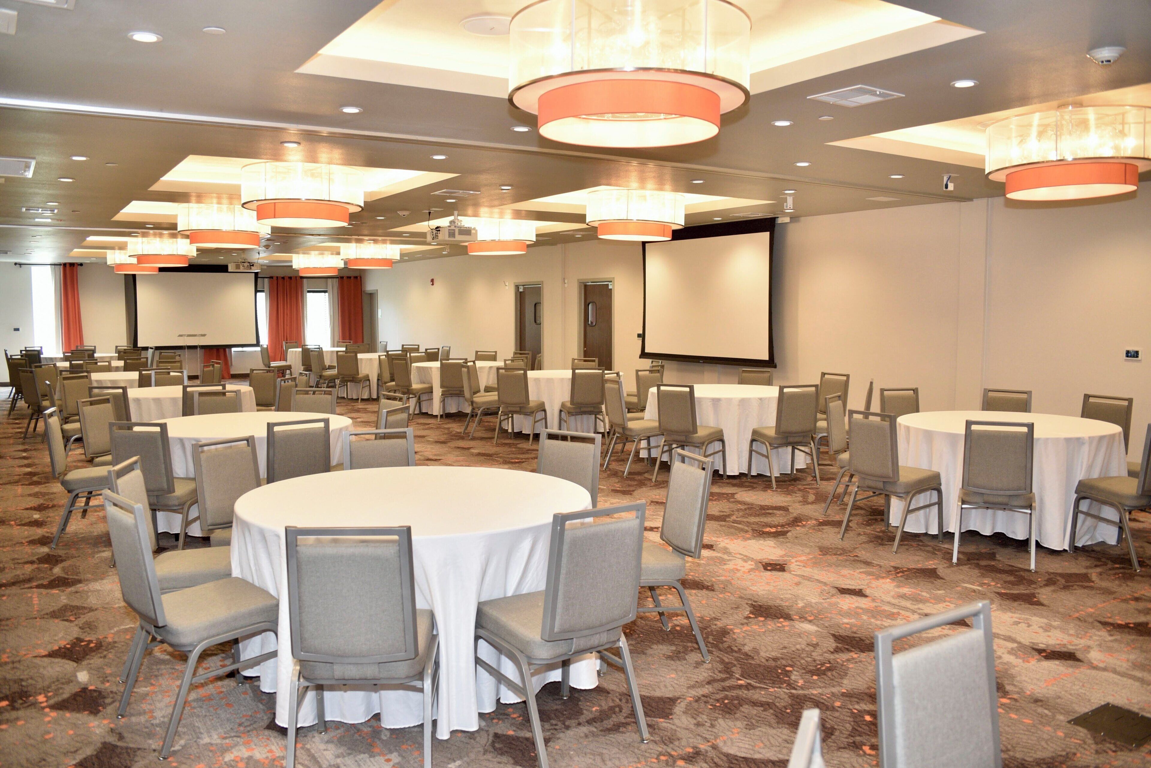 Sala de conferência com várias mesas e cadeiras, equipada com tela de projeção e iluminação embutida no teto.