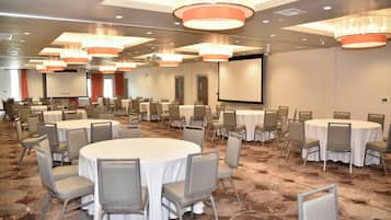 Sala de conferência com várias mesas e cadeiras, equipada com tela de projeção e iluminação embutida no teto.