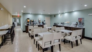 Uma área de refeições moderna com mesas e cadeiras brancas, um espaço para café e uma cozinha.