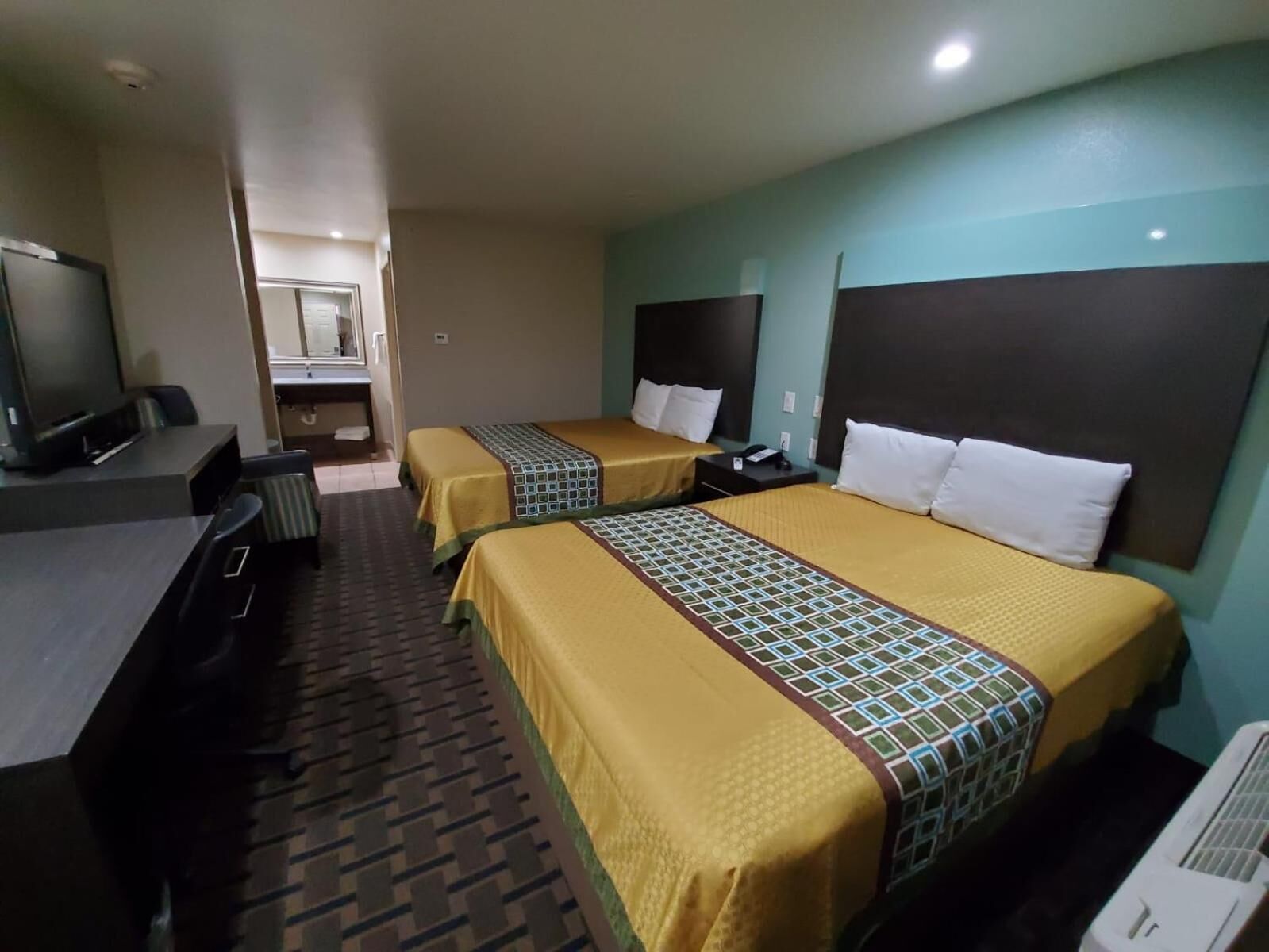 Quarto de hotel com duas camas, uma escrivaninha, televisão e banheiro visível por uma porta aberta.