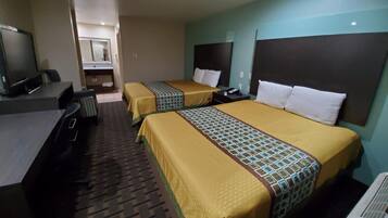Quarto de hotel com duas camas, uma escrivaninha, televisão e banheiro visível por uma porta aberta.