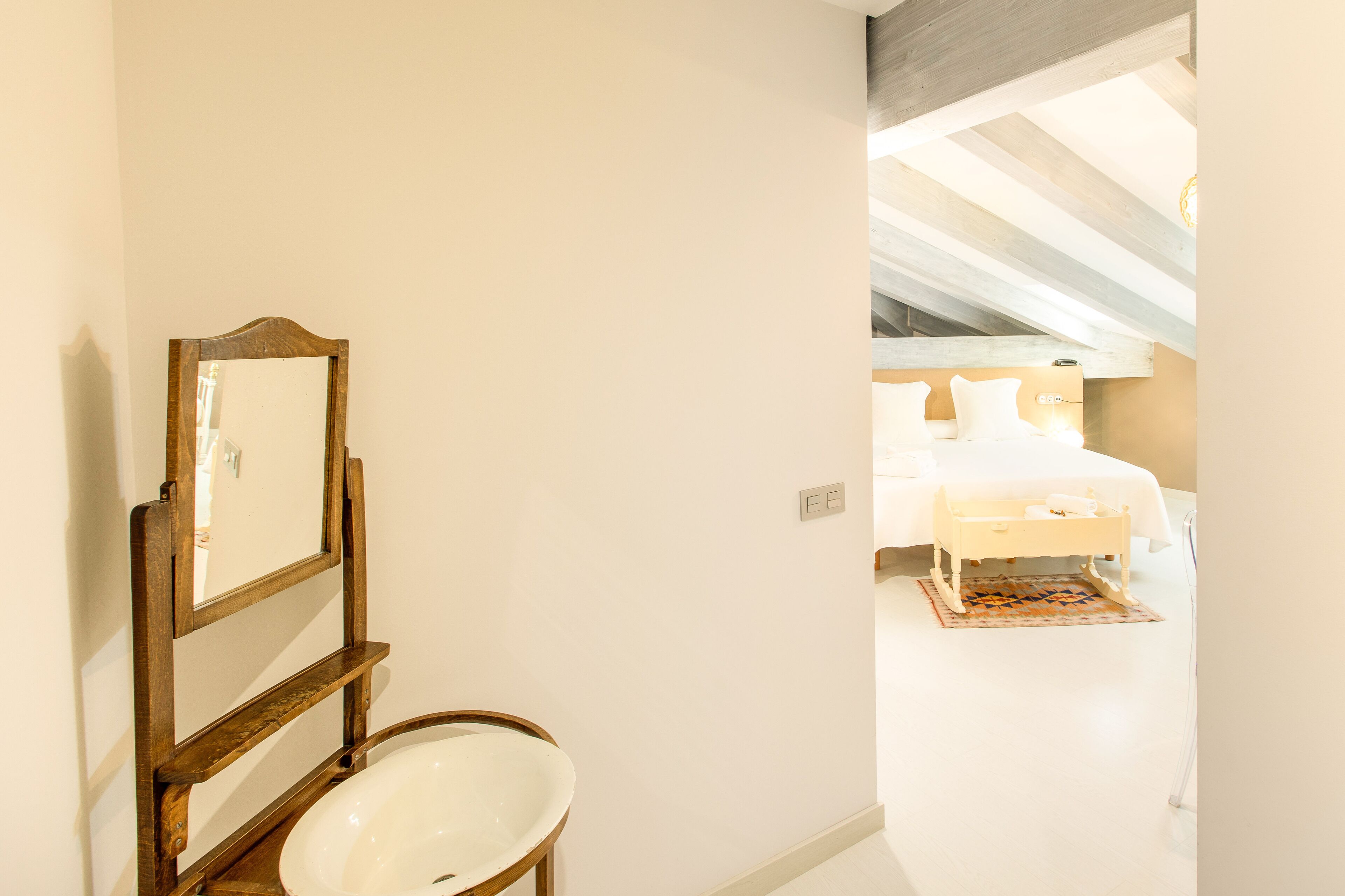 Casa de banho com espelho emoldurado a madeira, lavatório branco e cama com colcha branca ao fundo.