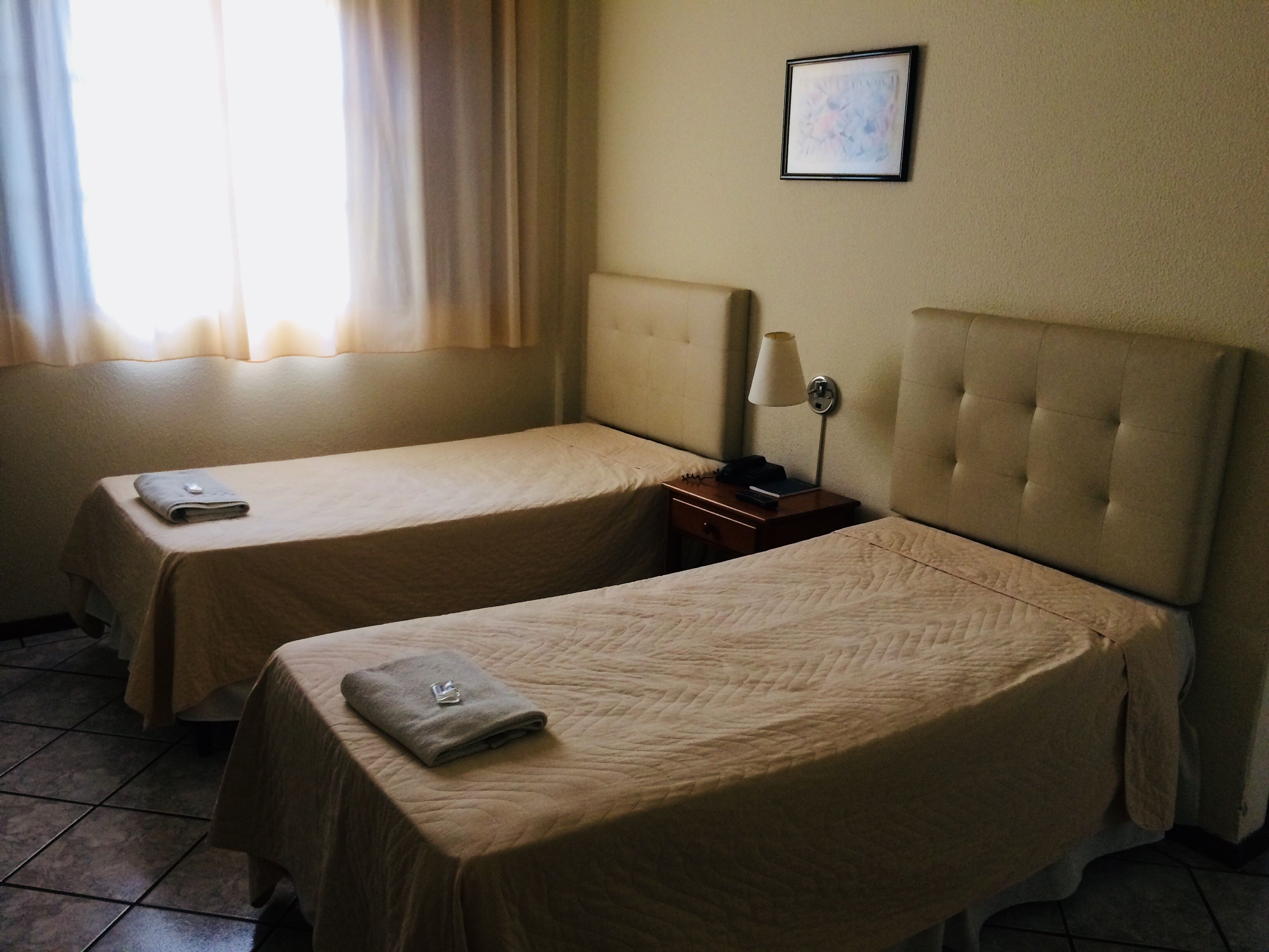 Quarto de hotel com duas camas de solteiro, uma mesa de cabeceira com abajur, um quadro na parede e uma janela com cortinas.