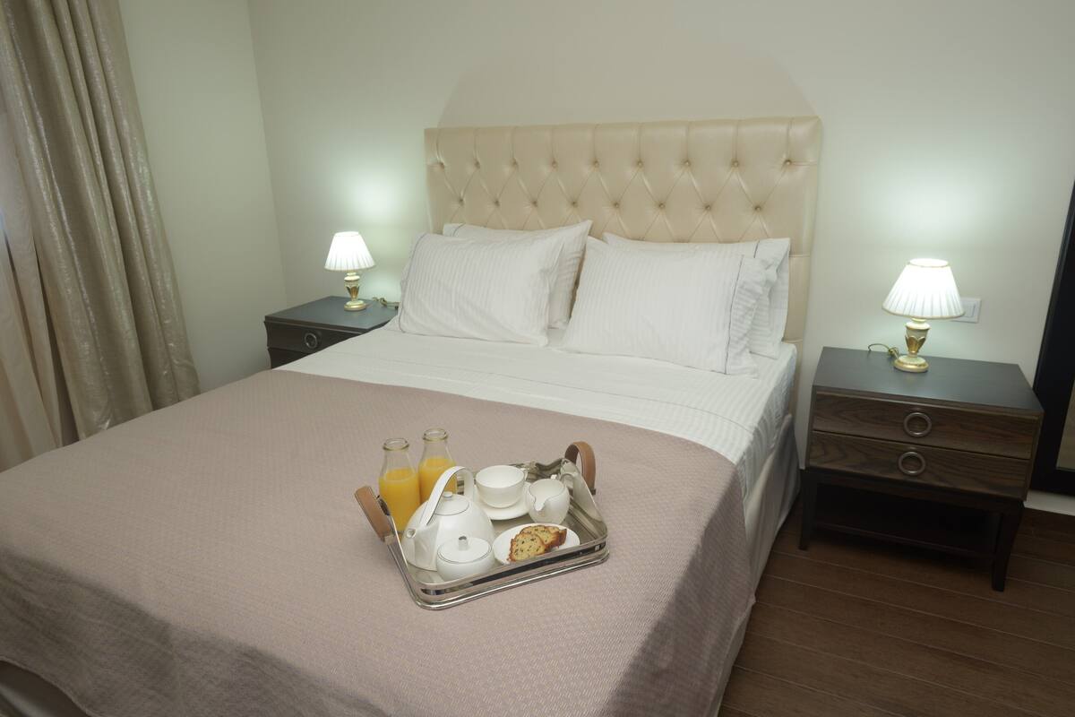 Cama bem arrumada com uma bandeja de itens de café da manhã, incluindo suco, um bule de chá e doces, em um quarto de hotel com cabeceira estofada e luminárias laterais.