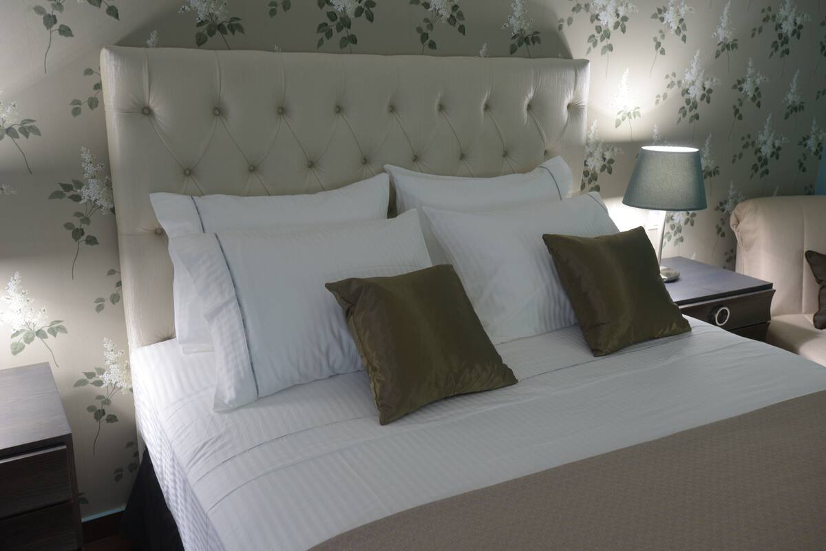 Cama bem arrumada com travesseiros brancos e castanhos, um cabeceira estofada e uma mesa de cabeceira com abajur.