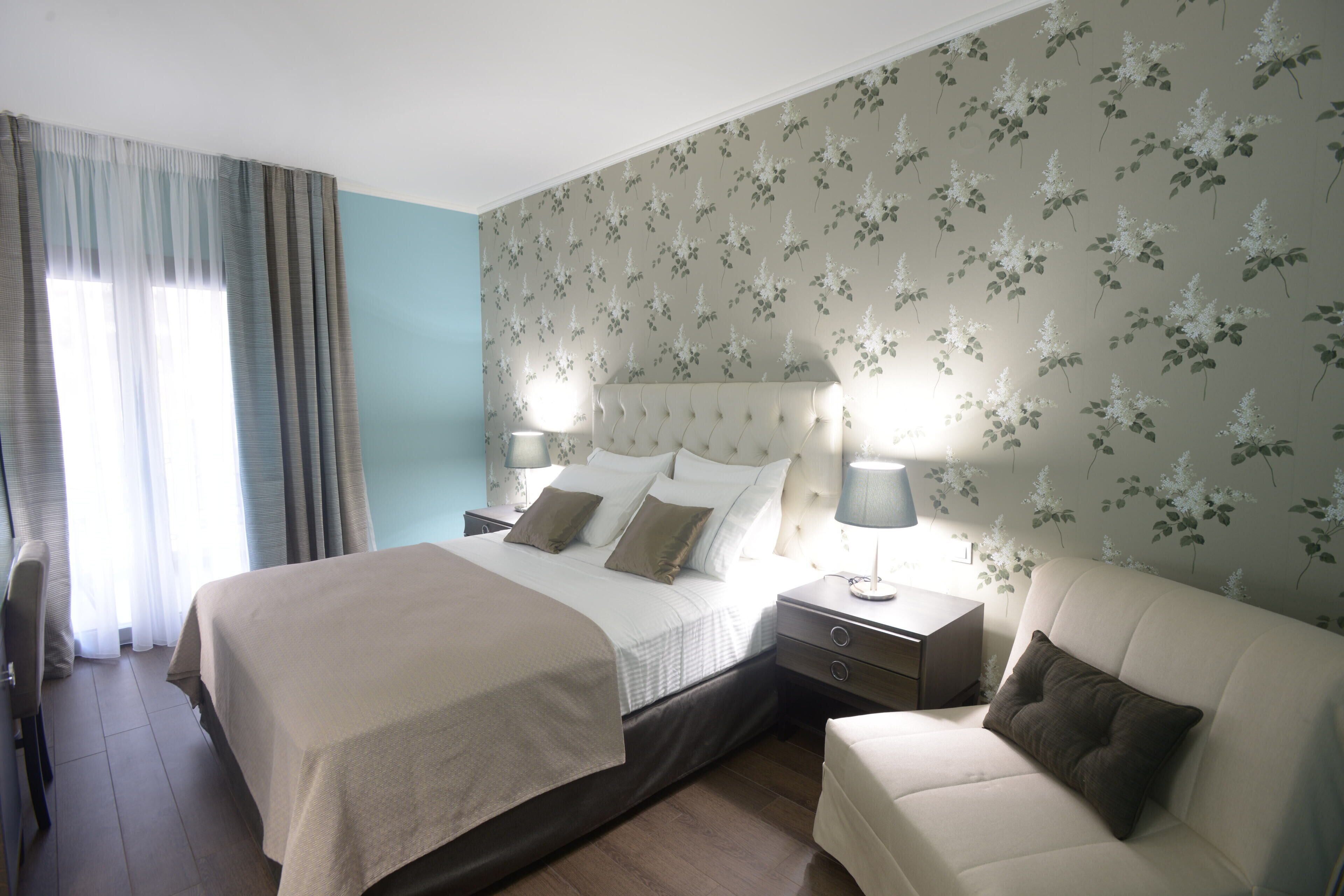 Quarto de hotel com uma cama grande, uma mesa de cabeceira com abajur, um sofá e papel de parede floral.