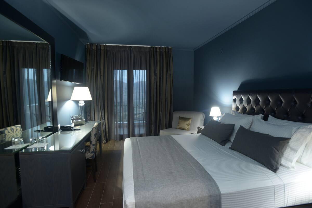 Quarto de hotel com uma cama grande, uma escrivaninha com luminária e vista para a cidade pelas janelas.
