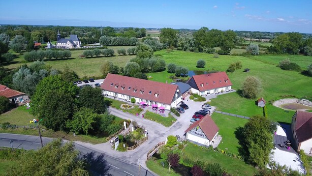 Vista aérea de uma propriedade extensa com vários edifícios, incluindo uma igreja, a casa principal e algumas construções menores. Há um estacionamento com carros, e a propriedade é cercada por vegetação exuberante e campos.