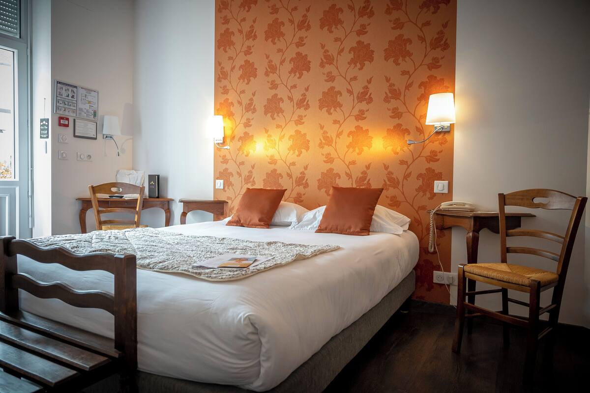 Cama bem arrumada com edredom branco e duas almofadas, uma cadeira de madeira com assento trançado, uma pequena mesa de madeira com abajur, e parede com papel de parede floral.