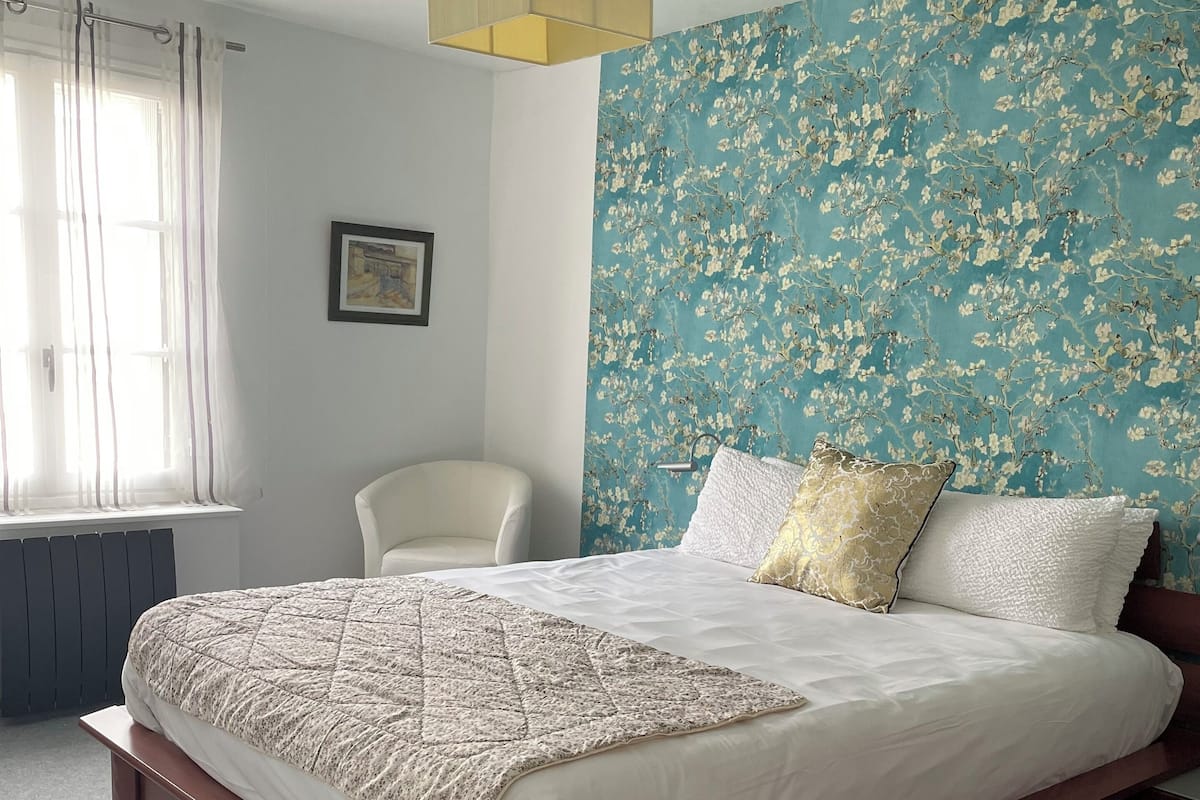 Quarto com cama de madeira, uma cadeira branca, janela com cortinas leves e papel de parede floral.