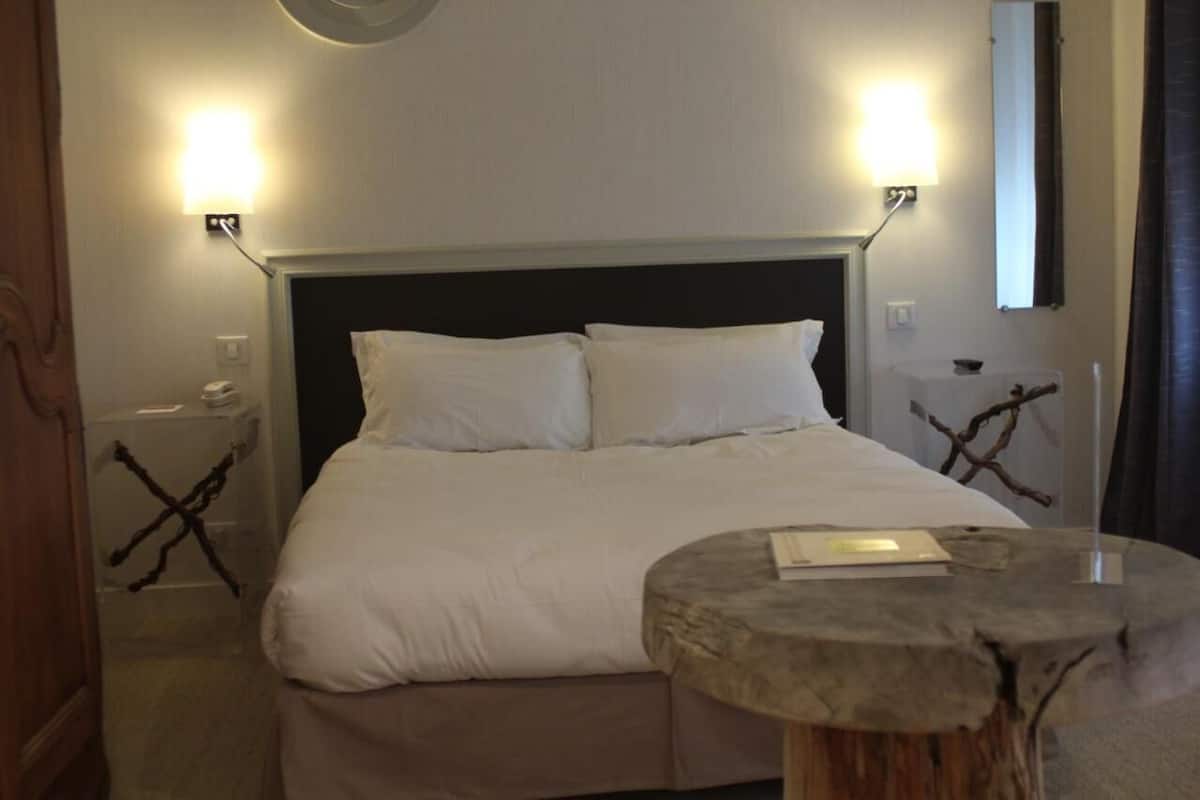 Uma cama com roupa de cama branca, uma mesa redonda de madeira e duas luzes fixadas na parede.