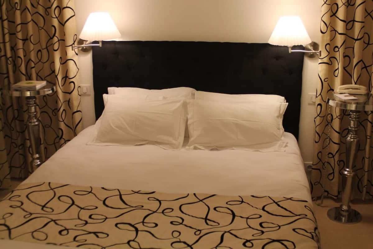 Uma cama com lençóis brancos, cabeceira preta e duas luminárias laterais.