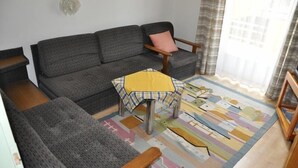 Uma sala de estar com um sofá cinzento, uma mesa pequena com toalha de mesa amarela e um tapete colorido.