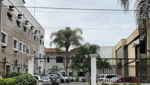 Uma área de estacionamento com vários carros, portão de acesso e edifícios com sacadas e aparelhos de ar condicionado.