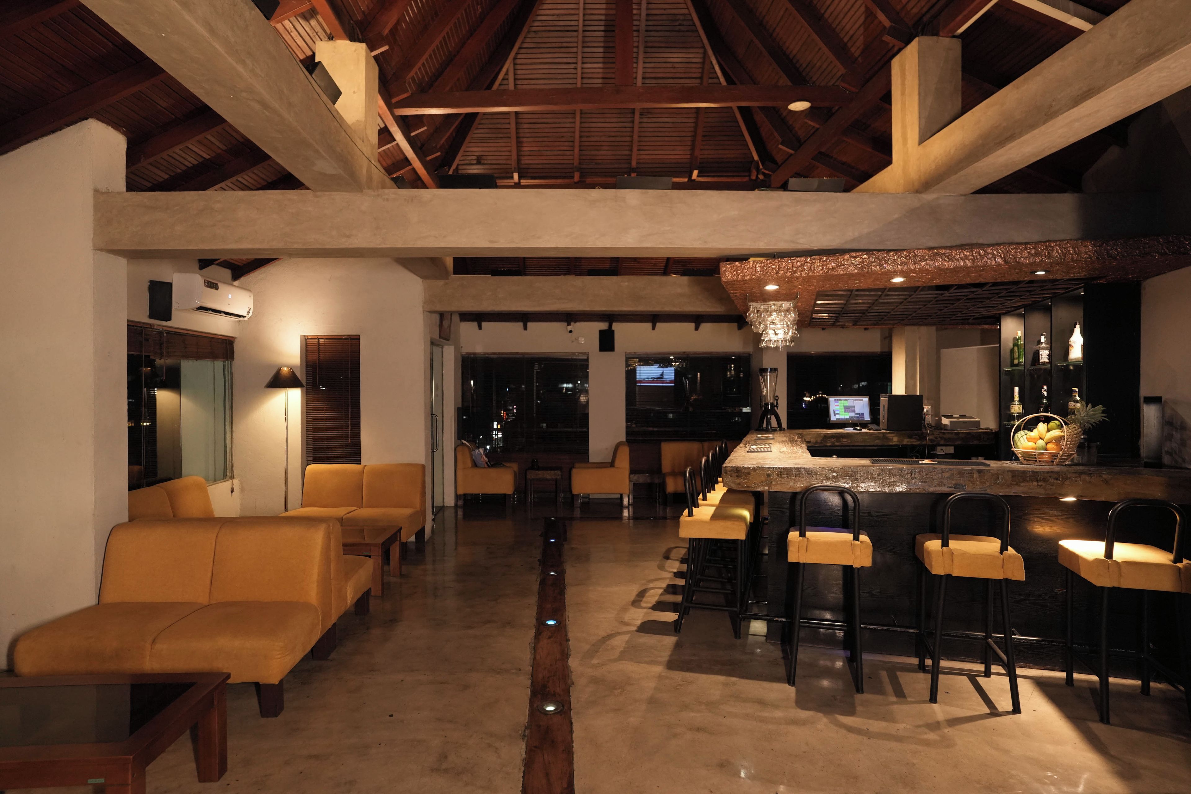 Amplo espaço de bar com pé-direito alto e vigas aparentes, destacando um balcão comprido com banquetas e uma tela de televisão.