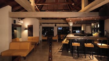 Amplo espaço de bar com pé-direito alto e vigas aparentes, destacando um balcão comprido com banquetas e uma tela de televisão.