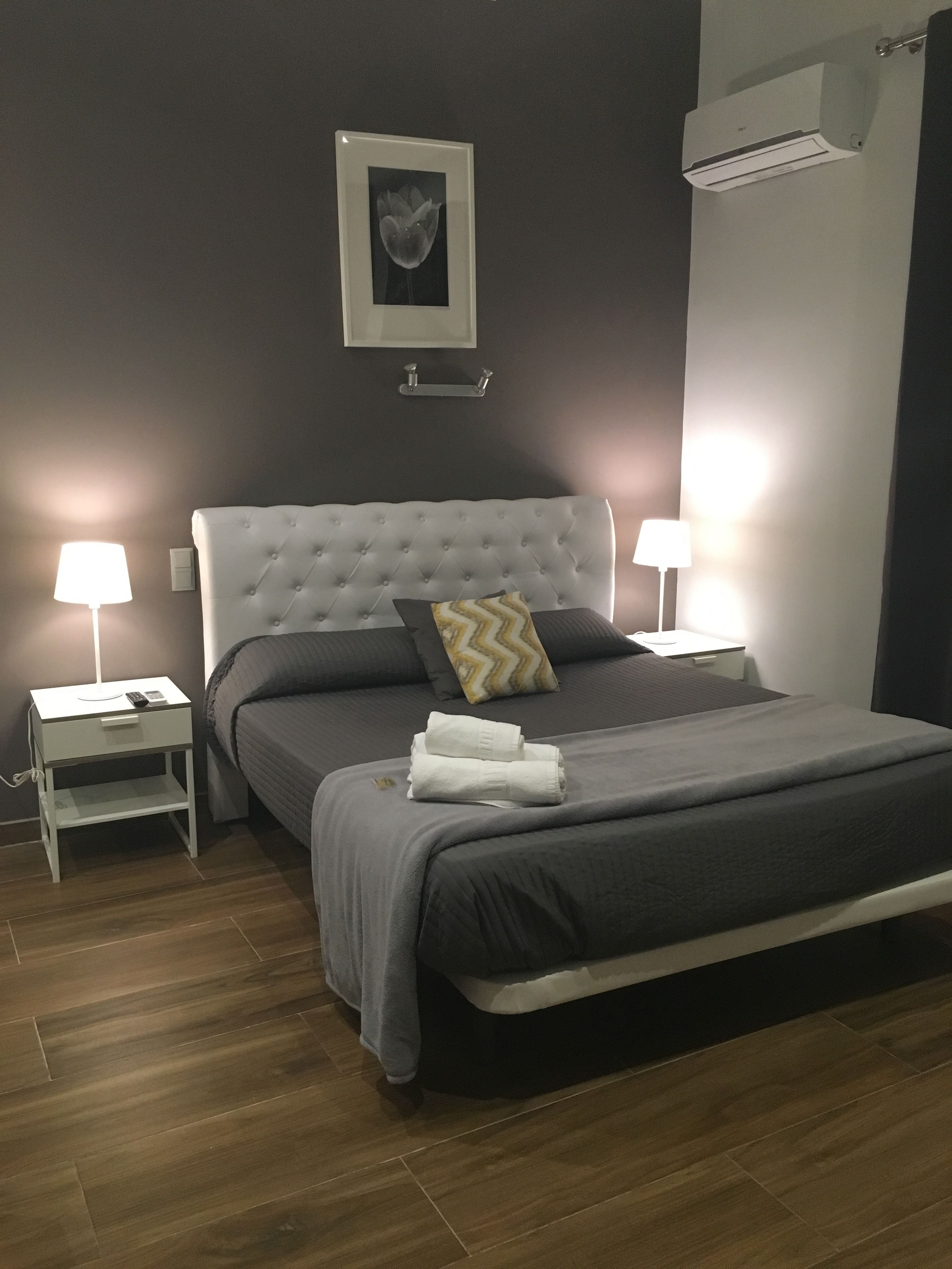Cama bem arrumada com cabeceira cinza, duas mesas de cabeceira com abajures, um quadro na parede e um aparelho de ar condicionado sobre a cama.