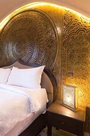 Uma cama com lençóis brancos, uma cabeceira escura e uma parede decorativa com padrões elaborados.