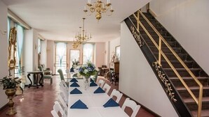 Uma sala de jantar formal com uma mesa comprida posta para refeição, uma escada com corrimãos dourados e um lustre.