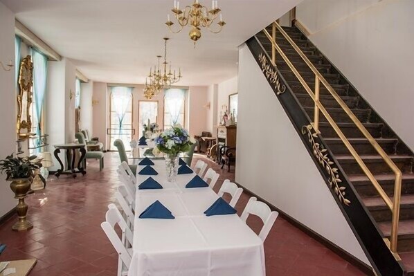 Uma sala de jantar formal com uma mesa comprida posta para refeição, uma escada com corrimãos dourados e um lustre.