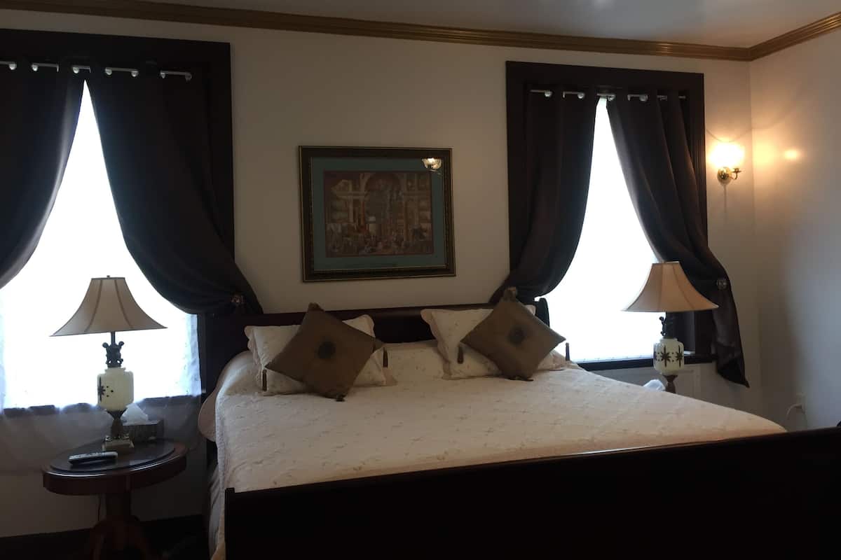 Quarto de hotel com uma cama grande, duas luminárias de cabeceira, um quadro na parede e cortinas escuras.