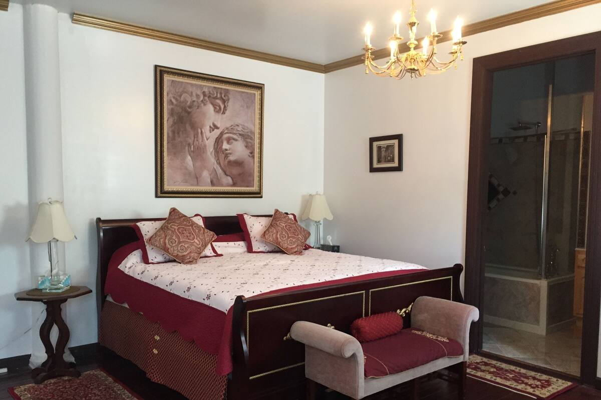 Quarto com uma cama grande, duas mesas de cabeceira, um candelabro, um quadro e uma porta de vidro deslizante para um banheiro.