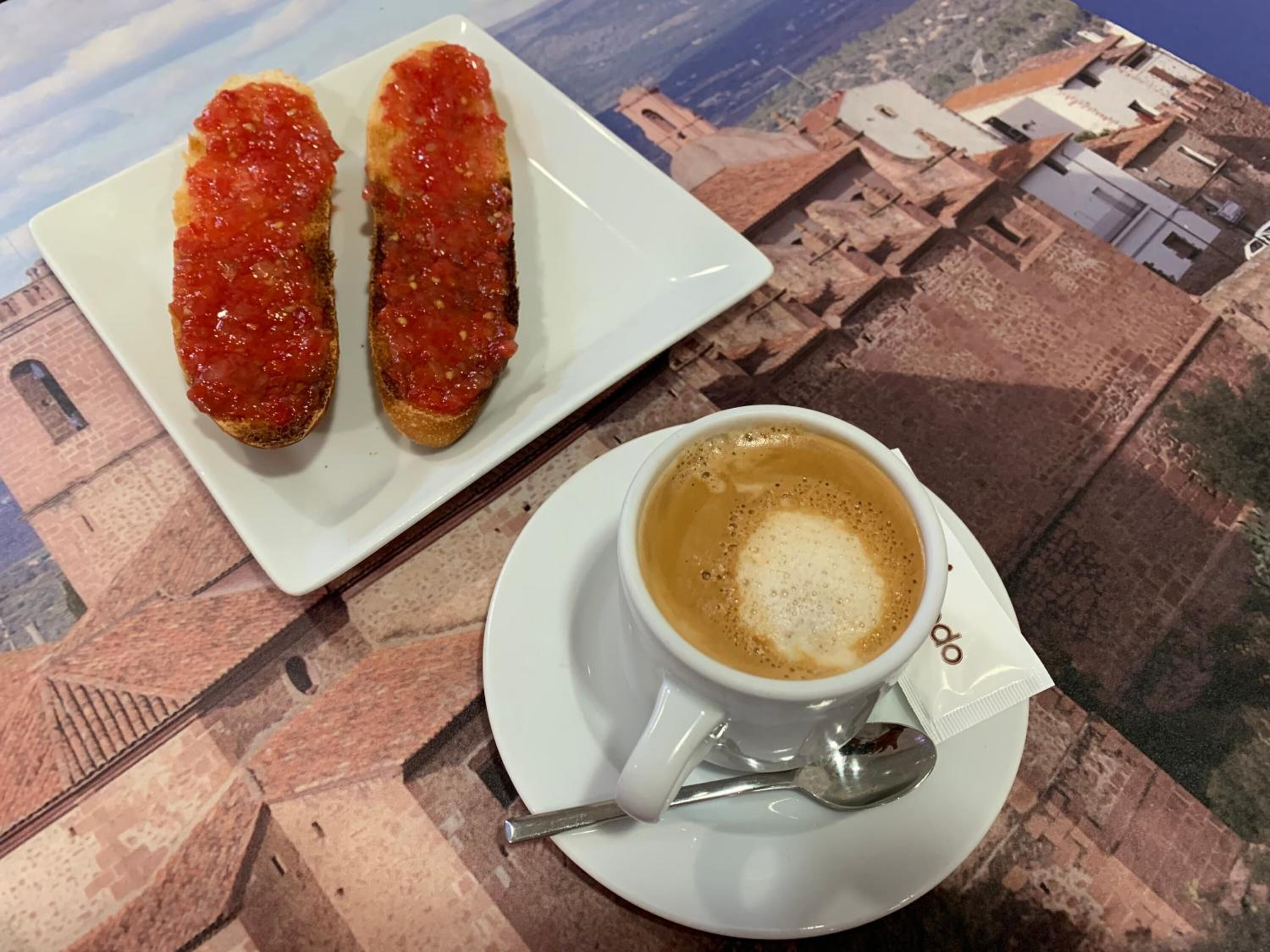 Um prato com duas fatias de pão cobertas com um creme vermelho e uma xícara de café com espuma sobre uma mesa com toalha estampada.