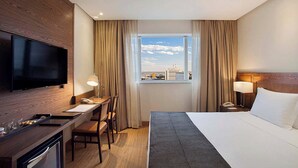 Quarto de hotel com cama, escrivaninha, cadeira, televisão e janela com vista para a cidade.