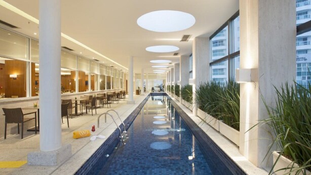 Piscina interior moderna com design longo e estreito, rodeada por áreas de estar e grandes janelas.