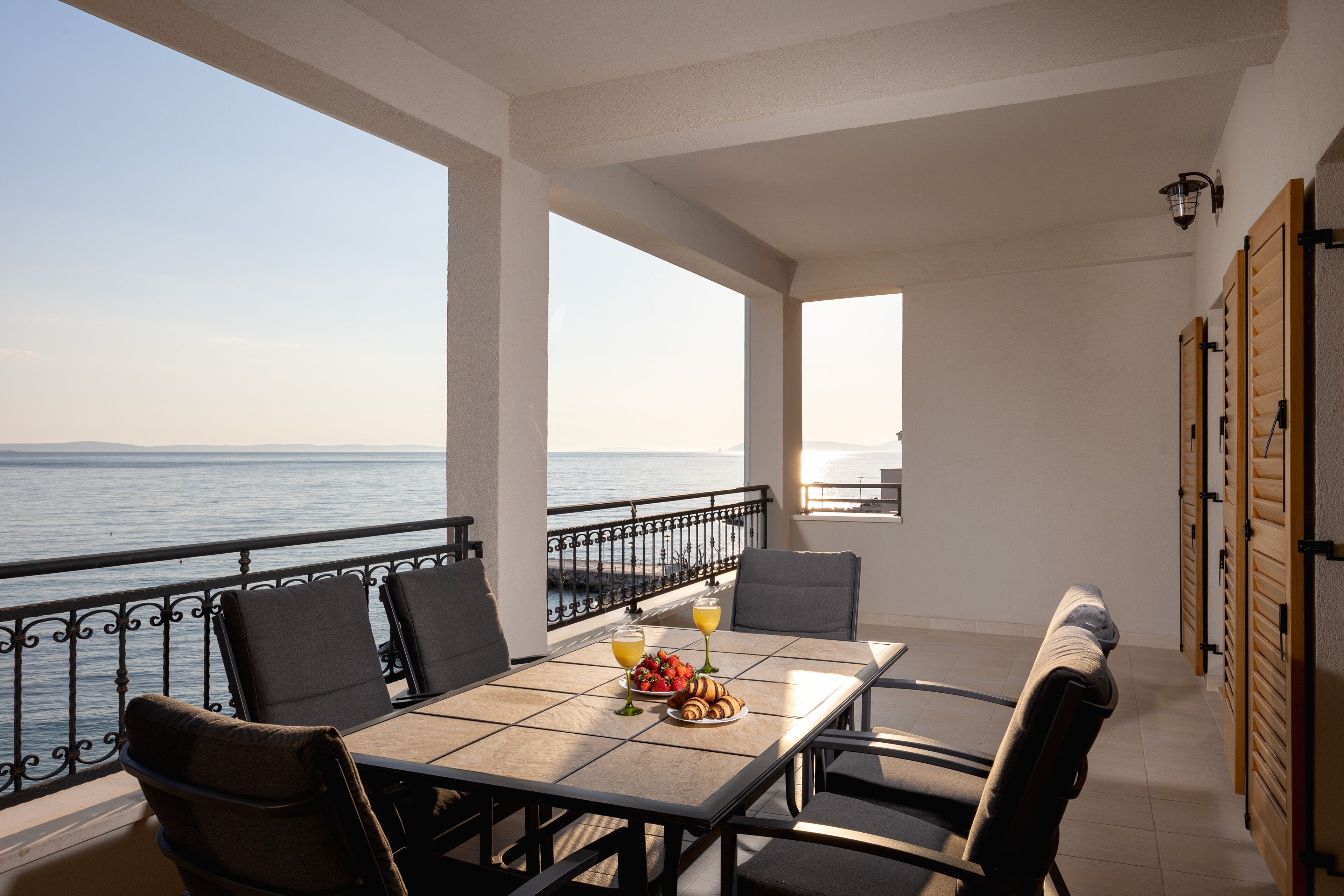 Uma varanda com uma mesa posta para dois, vista para o mar e uma fileira de portas de madeira.