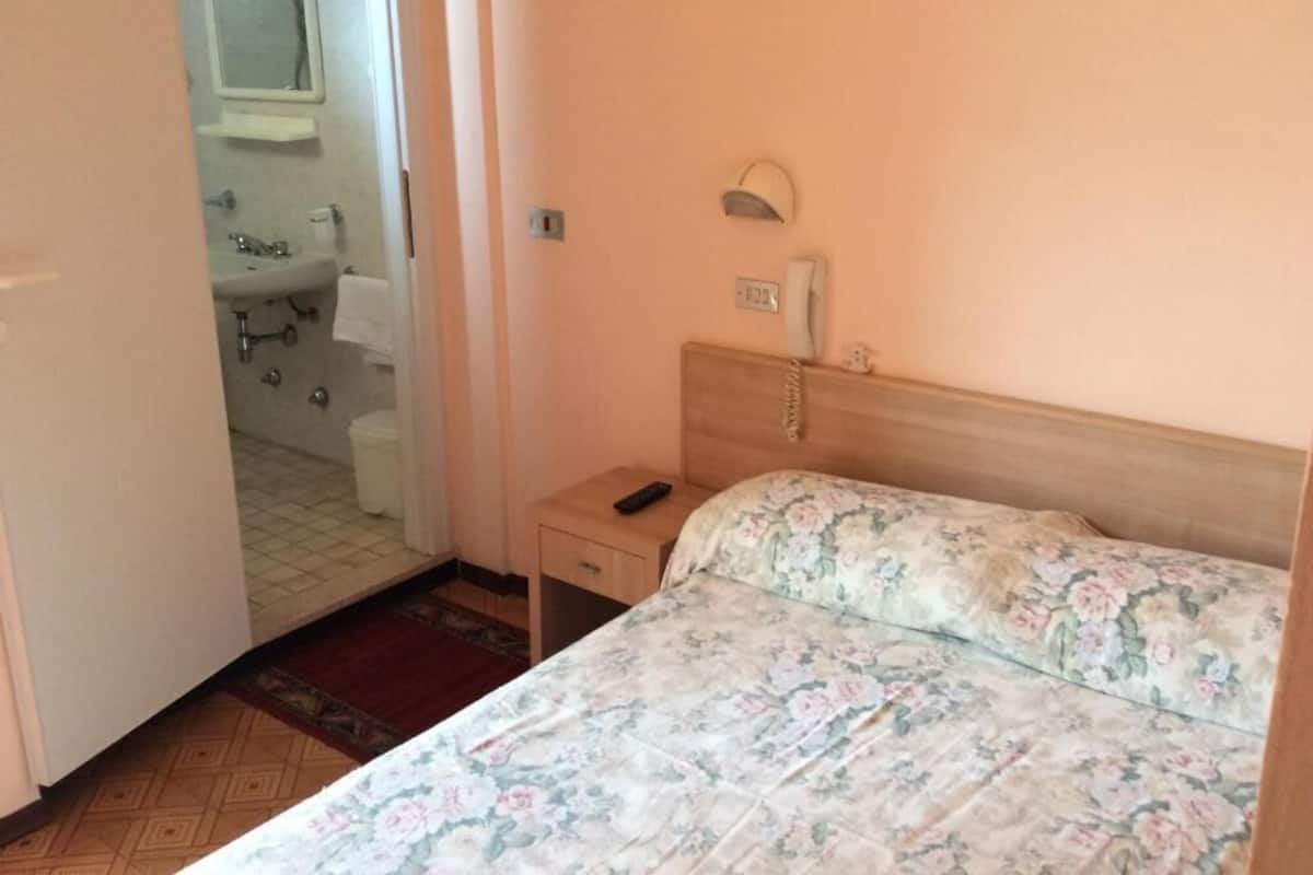 Quarto com cabeceira de madeira, uma cama de solteiro com roupa de cama florida, uma mesinha de cabeceira com controle remoto e um banheiro visível através de uma porta aberta.