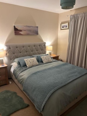 Quarto com cabeceira estofada, cama com colcha estampada em azul, duas luminárias de cabeceira, um quadro na parede e um tapete verde.