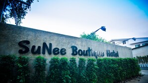 Placa do Hotel Boutique SuNee, com parede de concreto e folhagem verde.