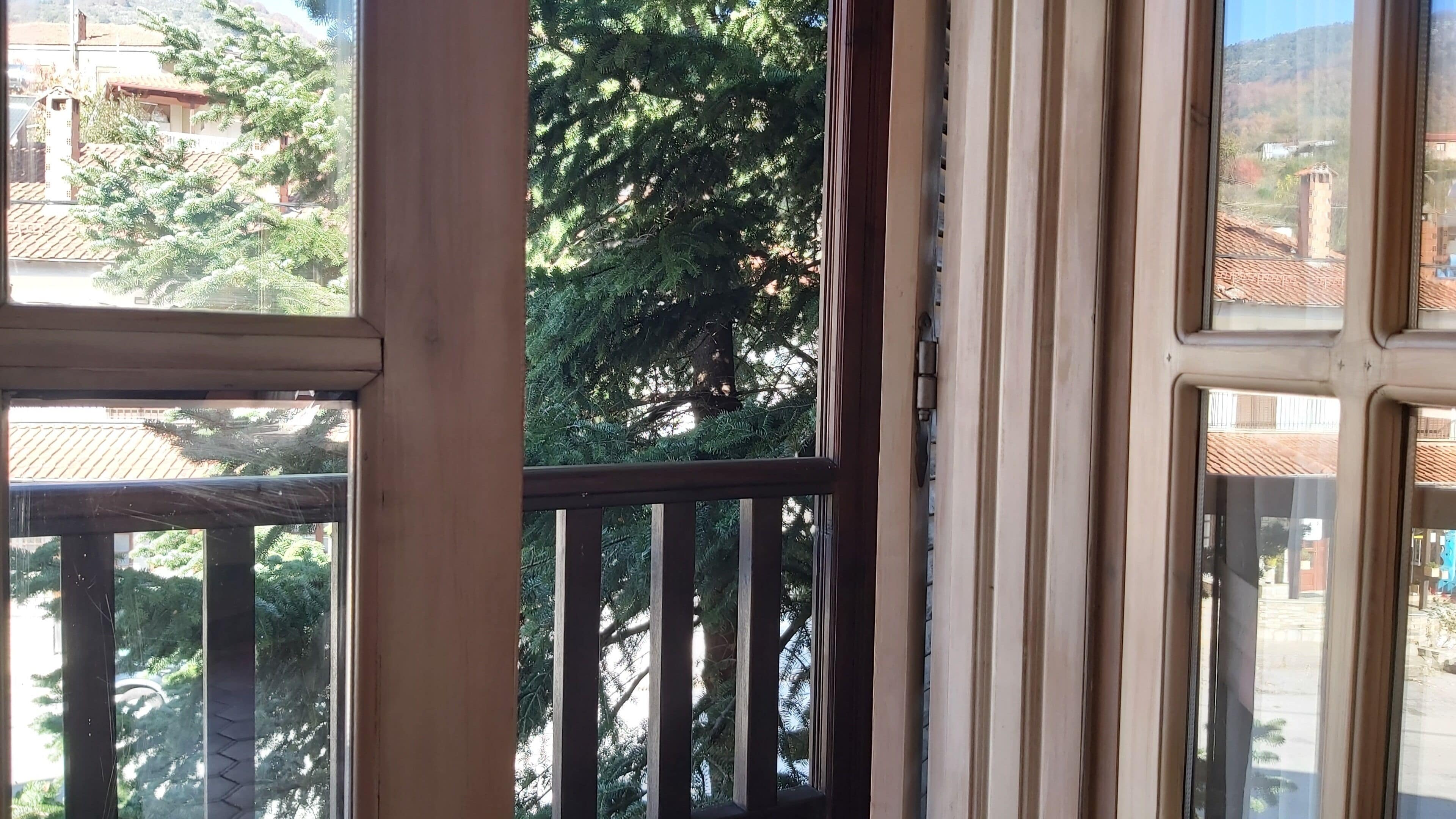 Vista pela janela, com moldura de madeira e corrimão metálico, sobre área residencial com casas e árvores.