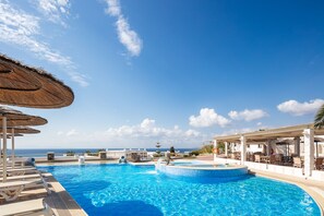 Piscina com espreguiçadeiras e guarda-sóis, área de estar coberta e vista para o oceano.
