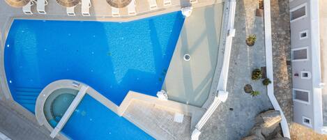 Vista aérea de uma piscina com várias áreas, rodeada por espreguiçadeiras e guarda-sóis, e adjacente a um edifício com fachada de pedra.